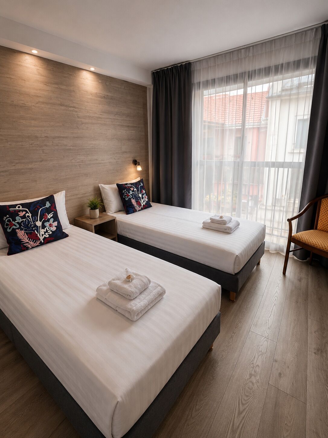 Quarto de hotel com duas camas, uma cabeceira de madeira, mesa de cabeceira com abajur, uma cadeira e uma janela grande com cortinas.