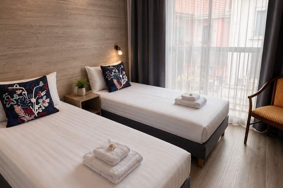 Quarto de hotel com duas camas, uma cabeceira de madeira, mesa de cabeceira com abajur, uma cadeira e uma janela grande com cortinas.