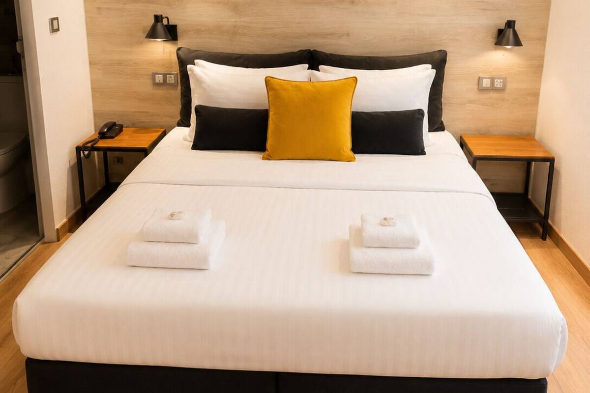 Cama bem arrumada com duas almofadas, mesa de cabeceira, abajur e luminária na parede.
