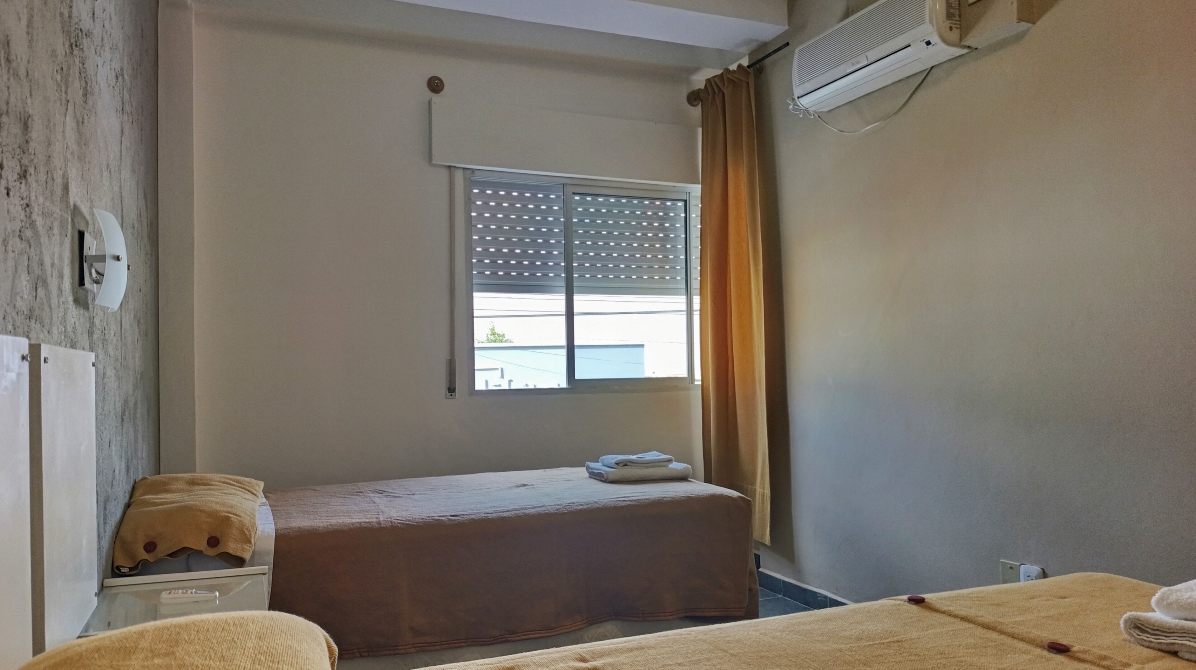 Quarto de hotel pequeno e simples, com uma cama, uma mesinha, uma janela com cortinas e um ar condicionado fixado na parede.