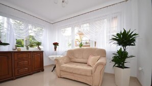 Quarto com sofá bege, armário de madeira, mesa branca com um vaso de flores e uma planta em vaso.