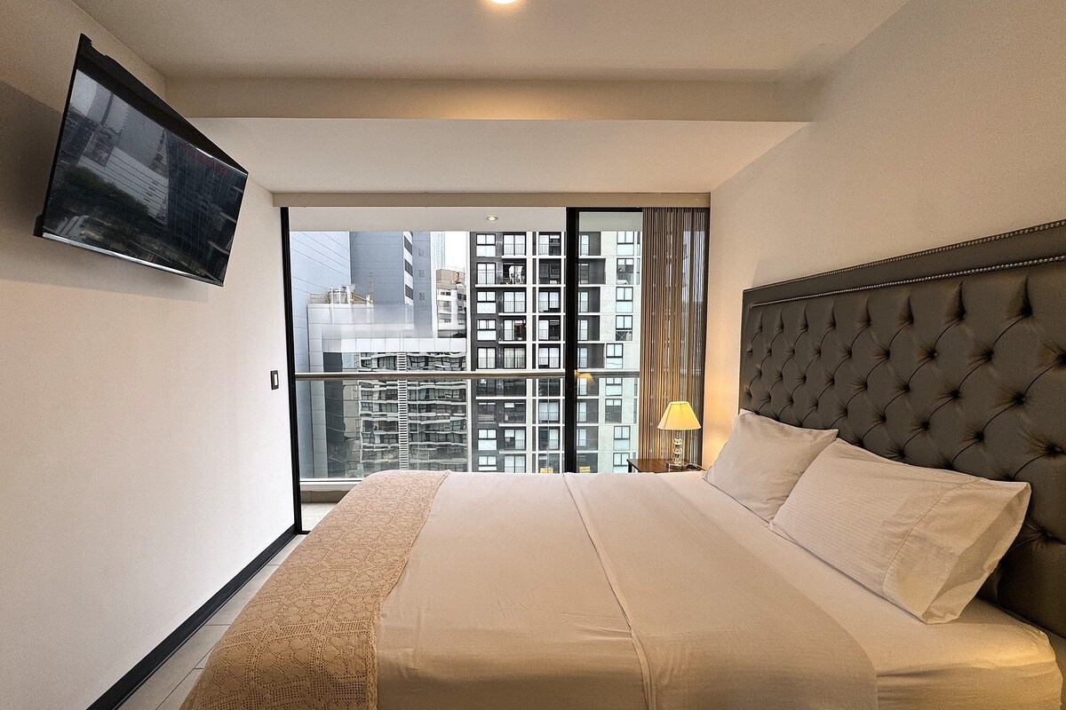Quarto de hotel com uma cama grande, televisão na parede e vista para edifícios altos pela janela.