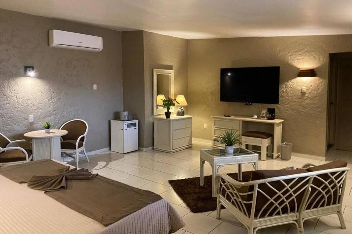 Quarto de hotel com cama, televisão, escrivaninha, cadeira, mesinha, luminária e ar condicionado.