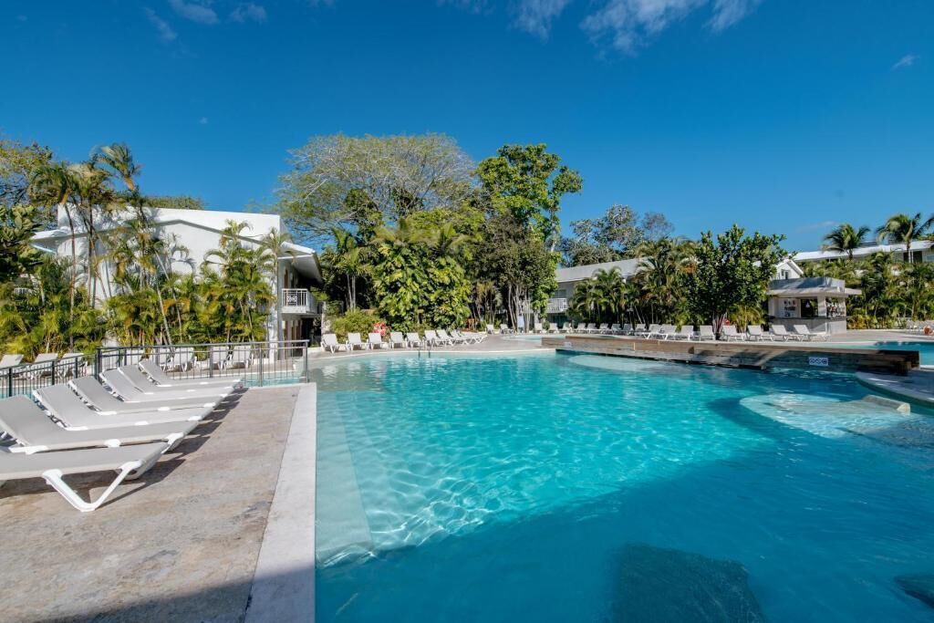 Uma piscina azul límpida com espreguiçadeiras, cercada por palmeiras e um edifício com varandas.