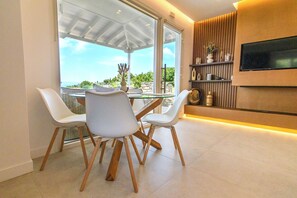 Uma zona de refeições moderna com mesa de vidro e cadeiras brancas, uma televisão na parede e vista para o exterior.