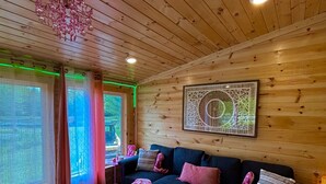 Um quarto com painéis de madeira, um sofá preto, um banquinho cor-de-rosa e uma peça decorativa na parede.