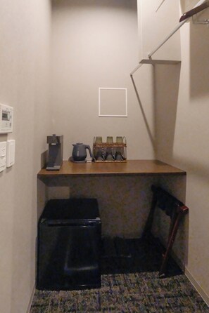 Uma pequena cozinha compacta com cafeteira, chaleira e micro-ondas. Há um cesto de lixo e uma cadeira no canto.