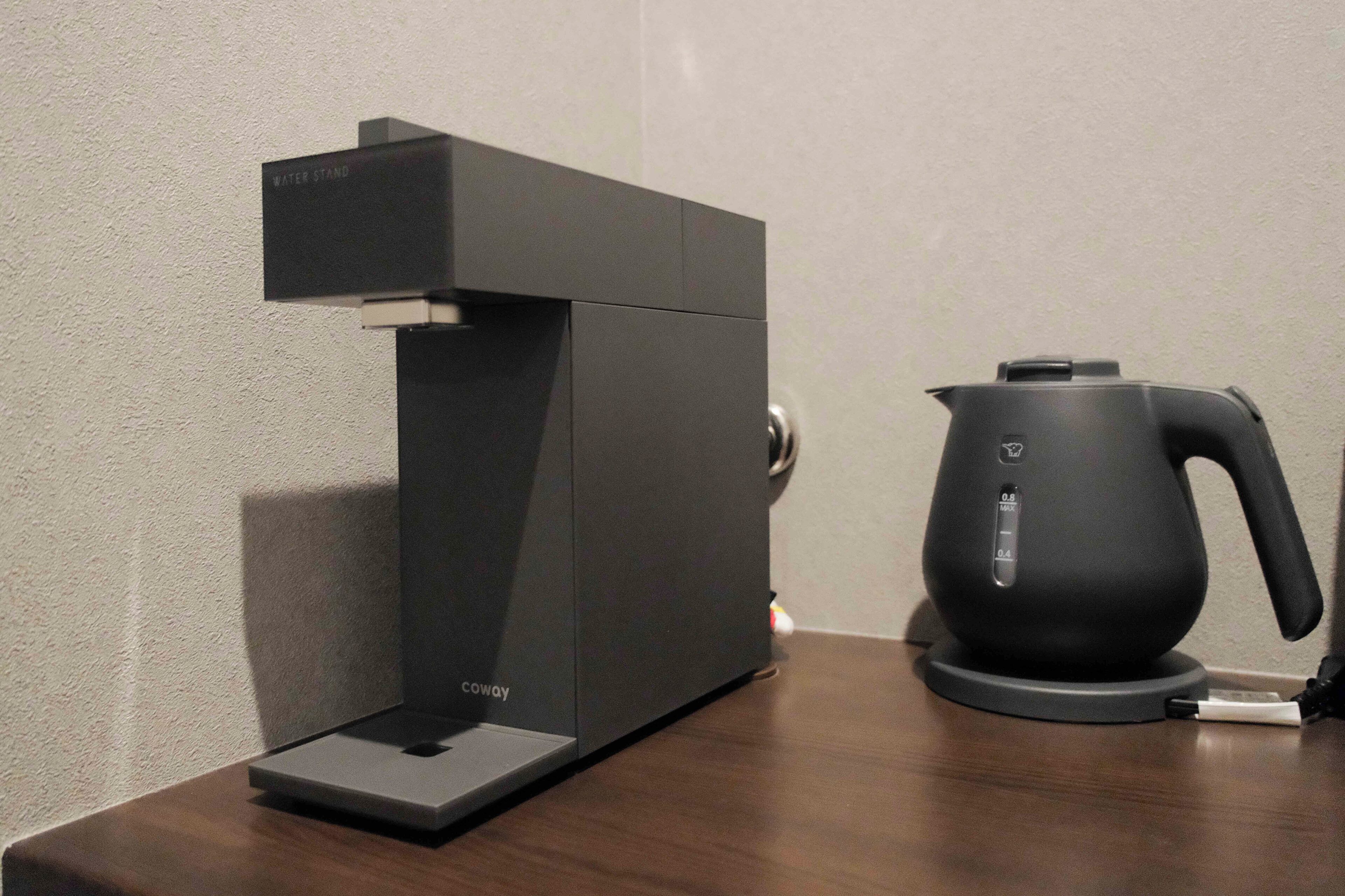 Uma cafeteira e uma chaleira sobre uma mesa de madeira em frente a uma parede lisa.