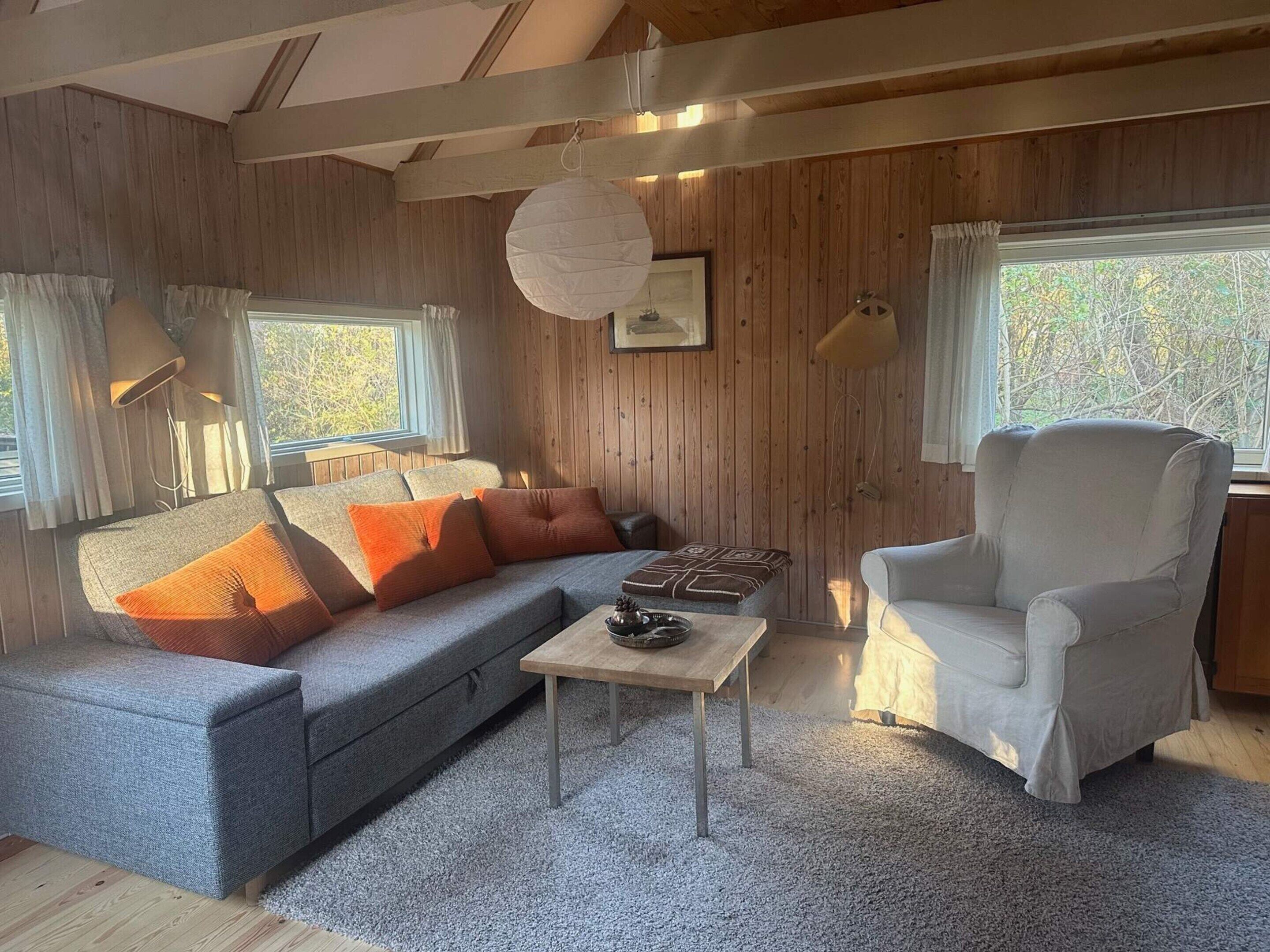 Um quarto acolhedor com um sofá cinzento, uma poltrona branca, uma mesinha e teto de madeira.