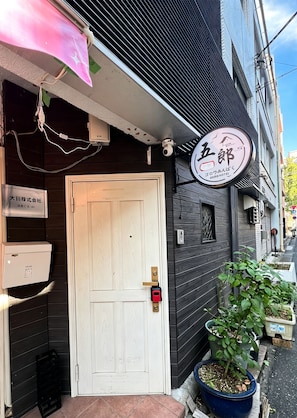 Entrada de hotel com porta branca, uma placa com caracteres japoneses e plantas em vasos do lado de fora.