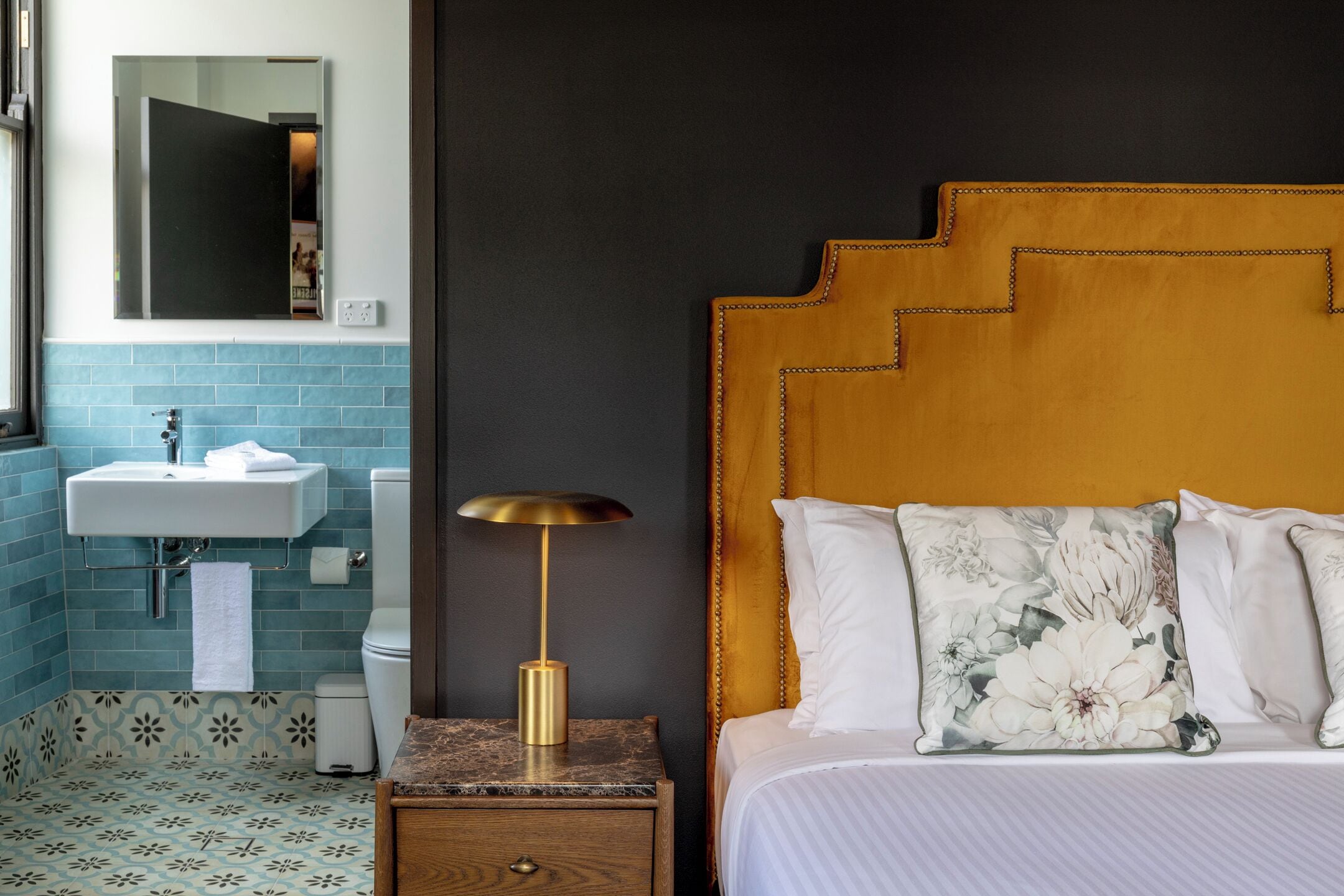 Quarto de hotel com banheiro, luminária de cabeceira, cama com almofadas estampadas e cabeceira.