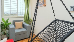 Um quarto com uma cadeira de rede preta, um sofá cinza com um travesseiro, uma mesa de madeira com um tecido vermelho e uma janela com cortinas verdes.