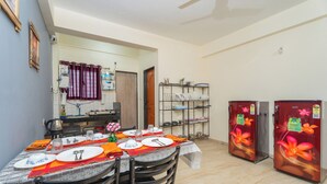 Uma área de refeições com uma mesa posta para quatro, cadeiras, uma cozinha com armários, uma geladeira e uma prateleira com louças.