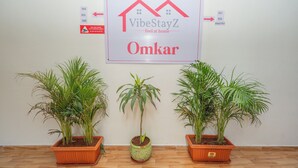 Placa indicando 'VibeStayZ Omkar' com setas mostrando a direção para os quartos 504, 505 e 506, além de uma placa para a despensa. Três plantas em vasos estão em frente à placa.