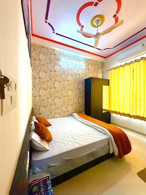 Quarto pequeno e bem iluminado com cama, guarda-roupa, ventoinha no teto e janela com cortinas.