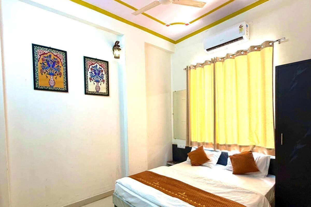 Quarto com cama, ventilador de teto, duas luminárias fixadas na parede, janela com cortinas e porta.