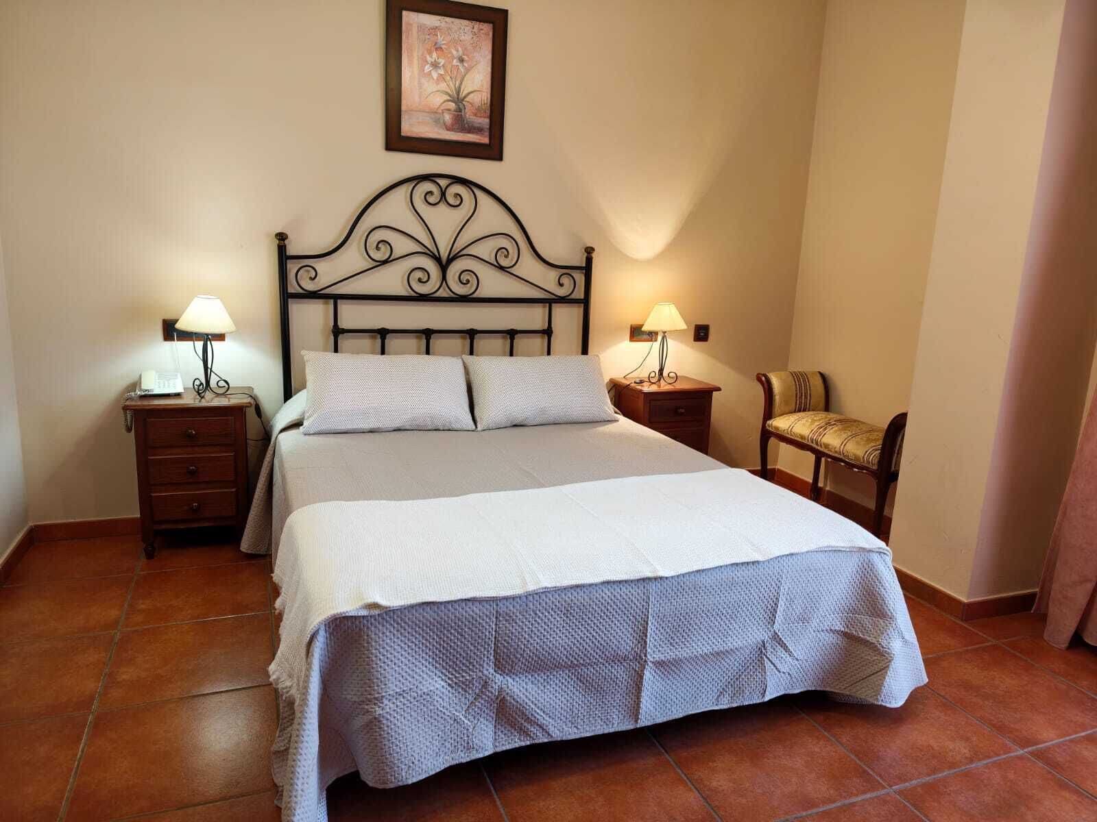 Uma cama bem arrumada com cabeceira, duas mesas de cabeceira com luminárias, uma cadeira e um quadro na parede.
