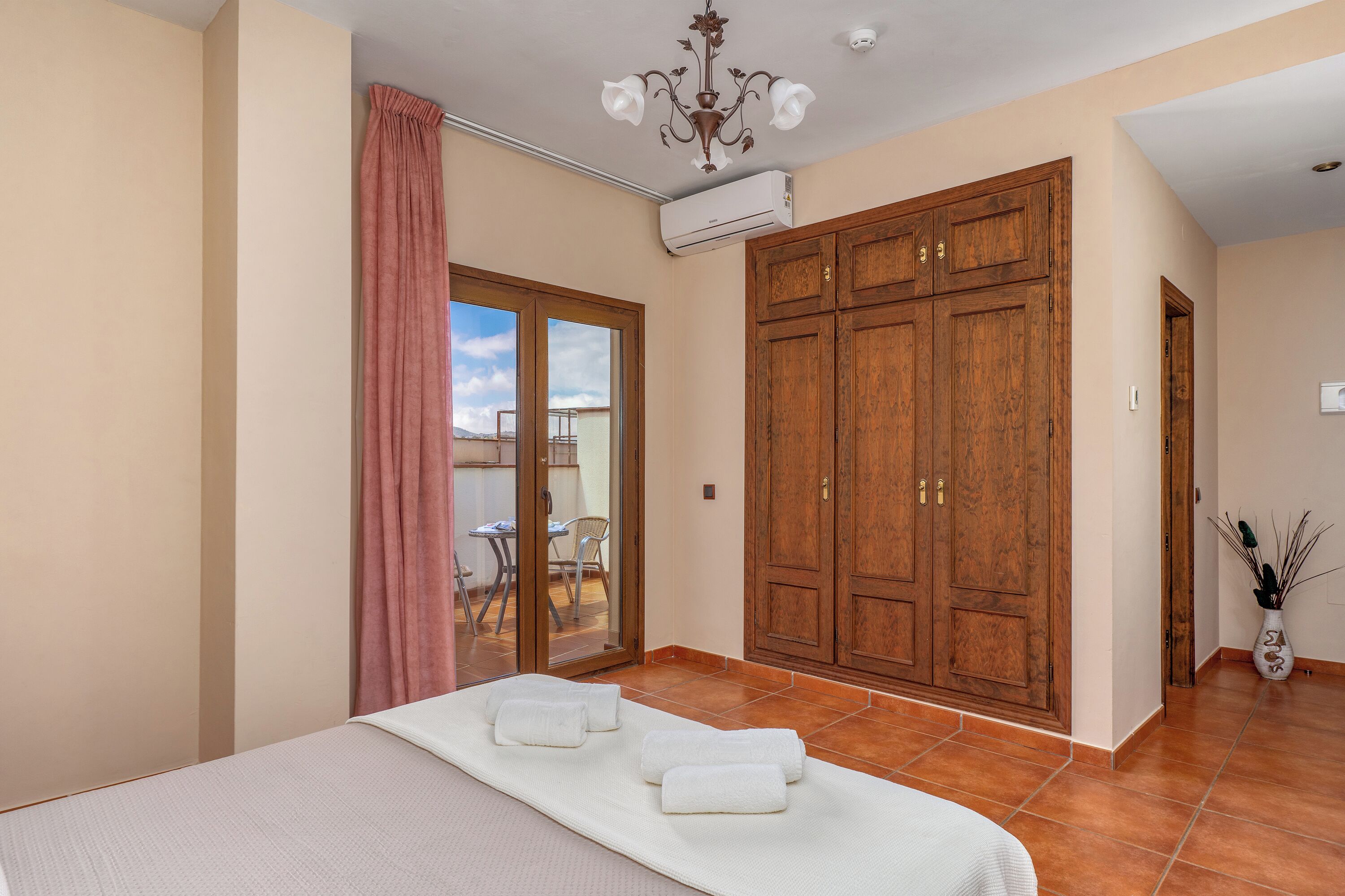 Um quarto com cama, guarda-roupa, varanda com cadeiras e uma luminária no teto.