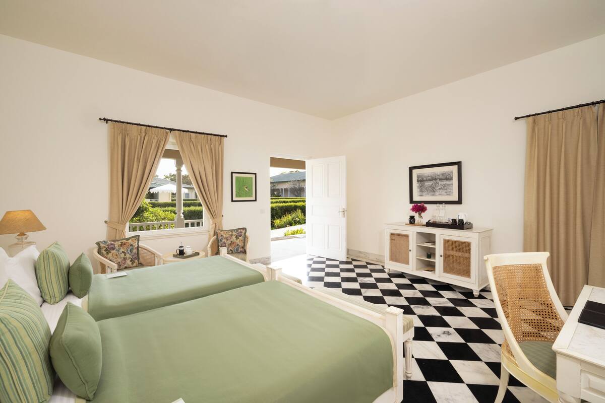 Quarto de hotel com duas camas, um chão xadrez, uma escrivaninha, uma cadeira e vista para o verde lá fora.
