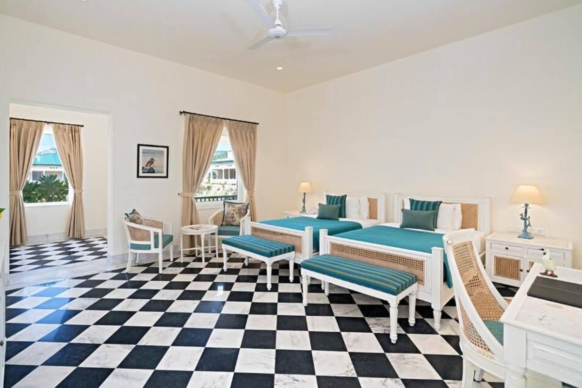 Quarto de hotel com chão quadriculado, cama com roupa de cama verde-água, escrivaninha com luminária e uma cadeira.