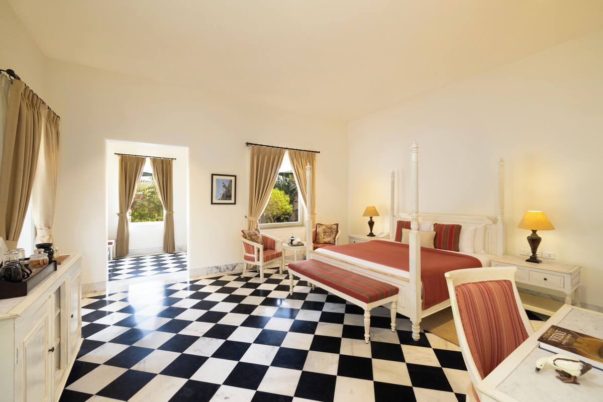 Quarto de hotel com chão xadrez, cama com colcha vermelha, uma cadeira, uma mesa pequena e um abajur.