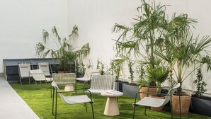 Uma área externa com assentos de vime, cadeiras e uma mesa redonda, cercada por plantas em vasos e uma parede branca.