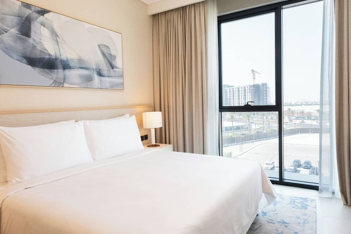 Quarto de hotel com uma cama grande, uma mesa de cabeceira com abajur, uma janela com vista para a cidade e um quadro na parede.