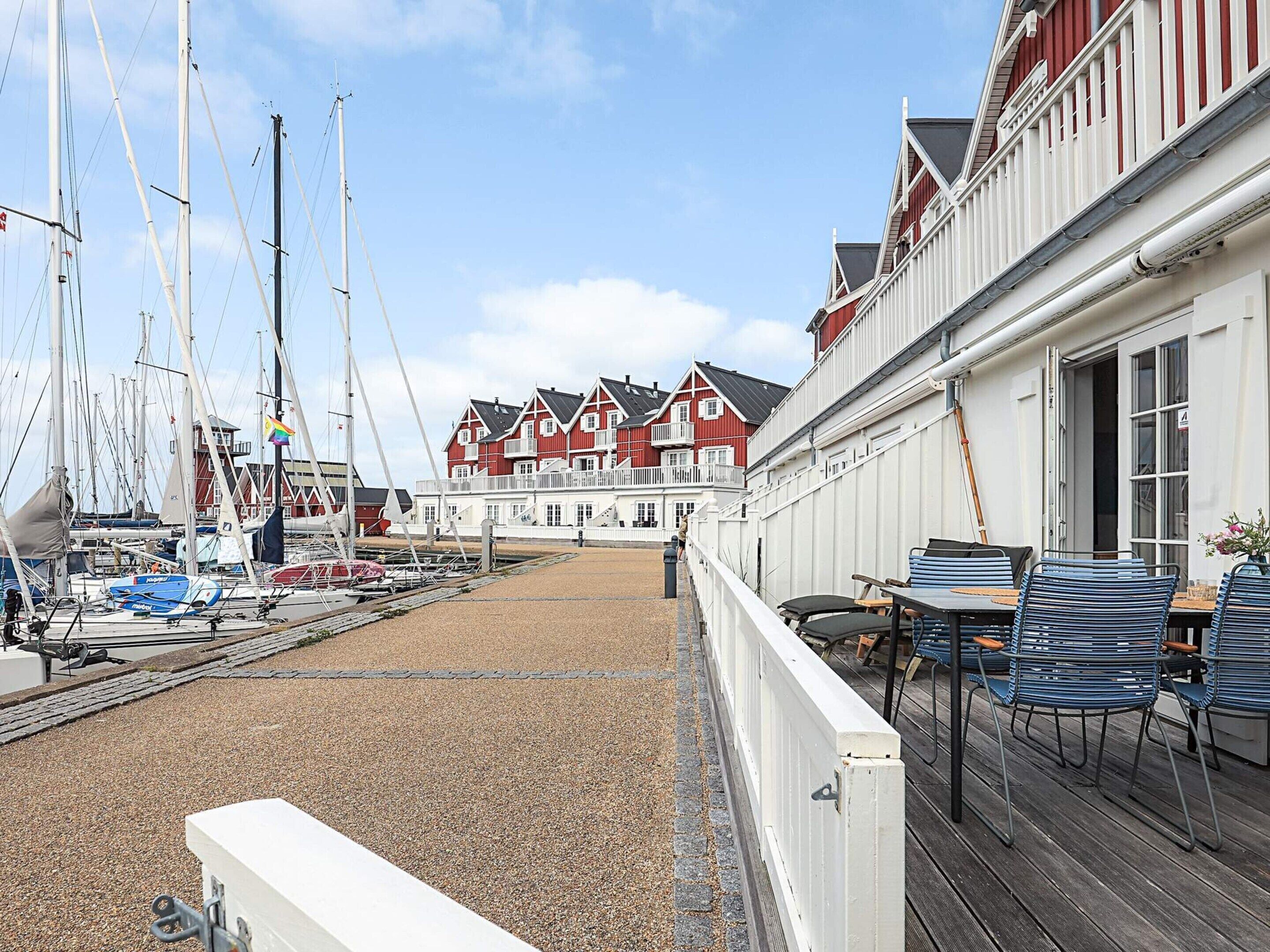 Marina com barcos atracados, um deck de madeira com cadeiras e um edifício com fachada vermelha e branca.