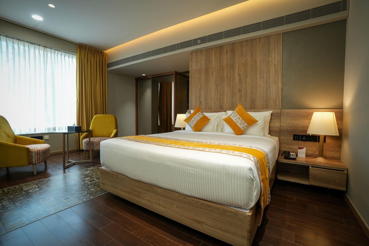 Quarto de hotel com uma cama grande, mesa de cabeceira com abajur, uma cadeira e escrivaninha com telefone.