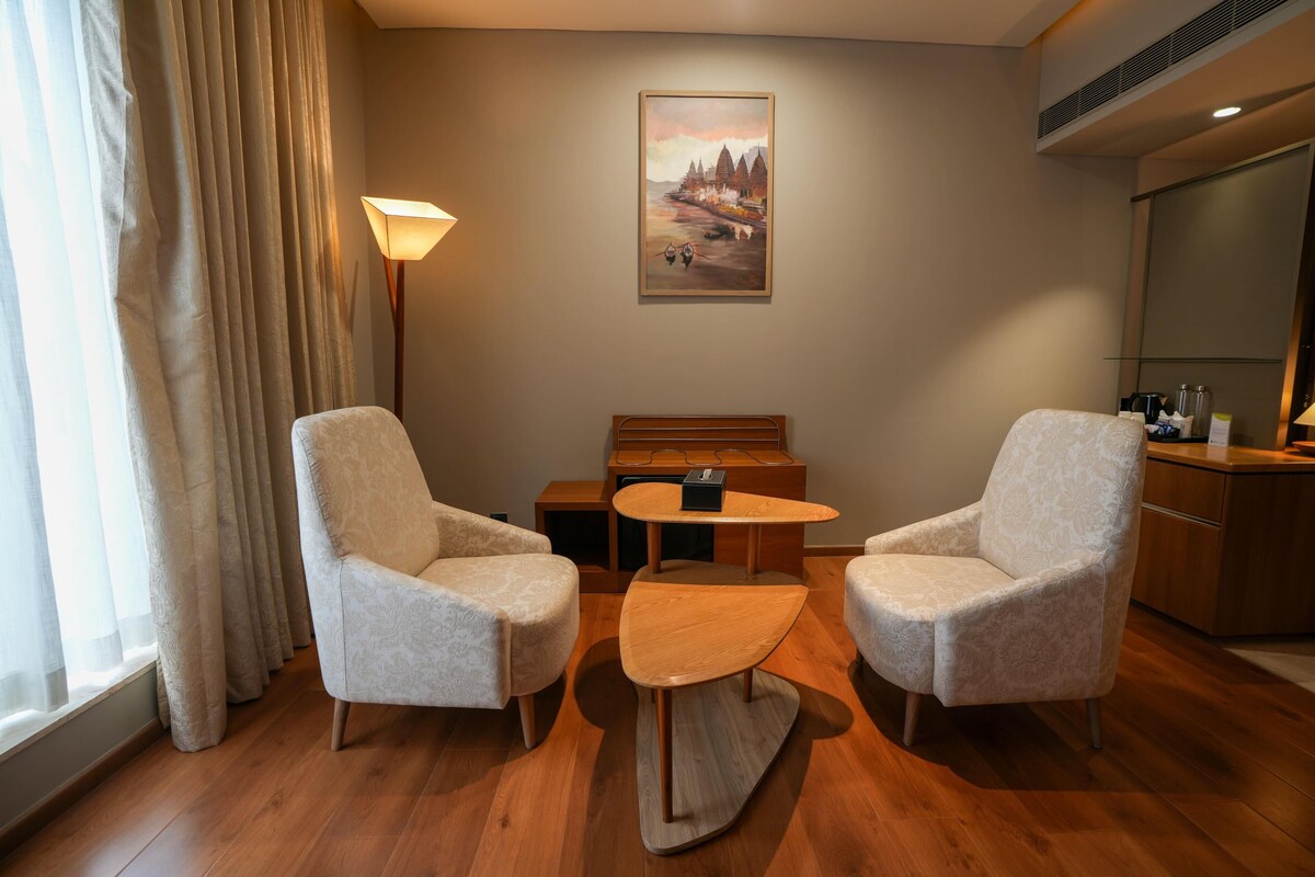 Quarto de hotel com duas poltronas, uma pequena mesa de madeira, um sofá, um quadro na parede e uma área de cozinha compacta.