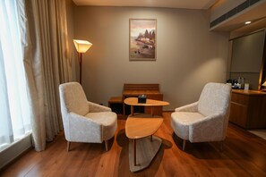 Quarto de hotel com duas poltronas, uma pequena mesa de madeira, um sofá, um quadro na parede e uma área de cozinha compacta.