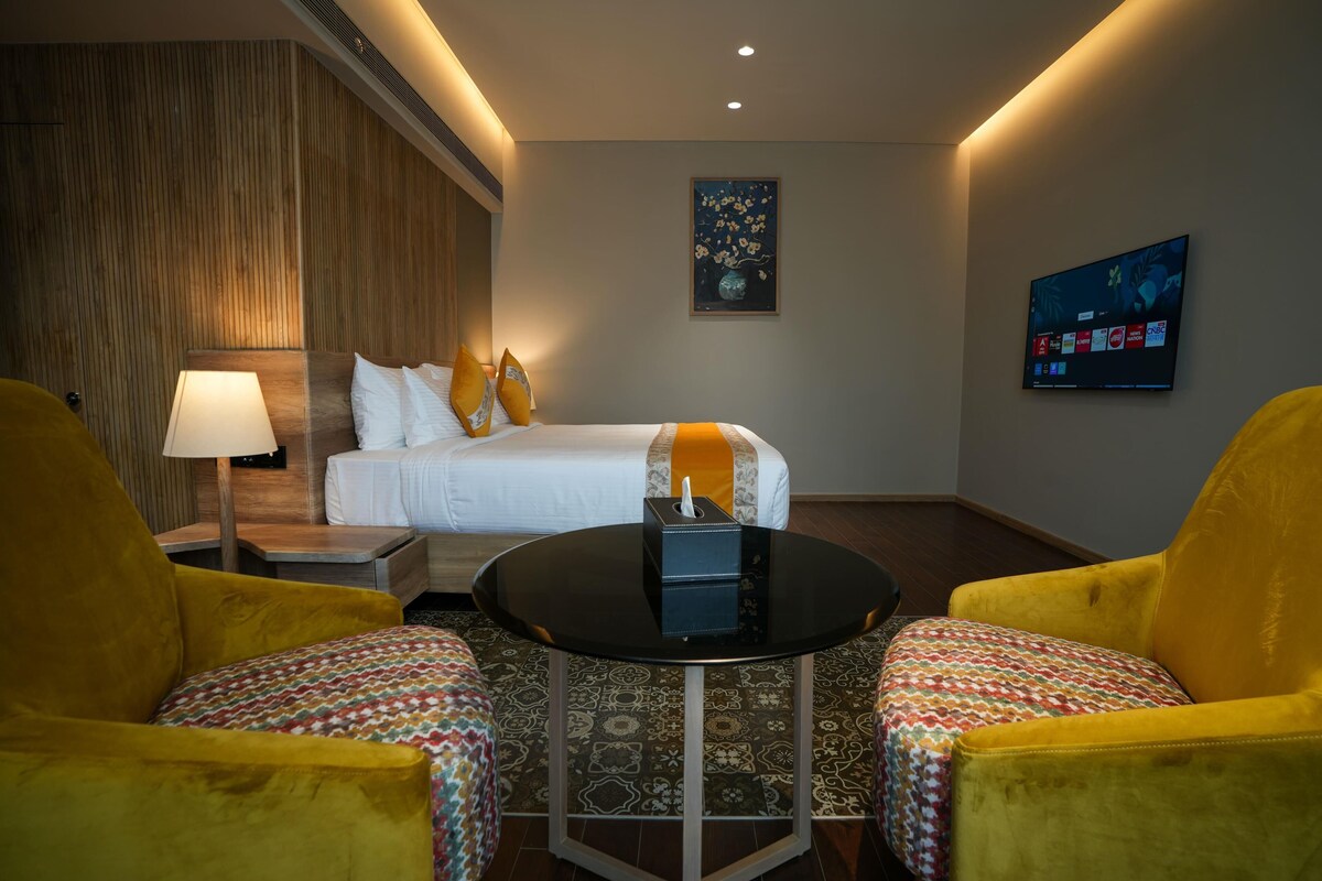 Quarto de hotel com cama, mesa de cabeceira com abajur, televisão na parede e duas poltronas amarelas.