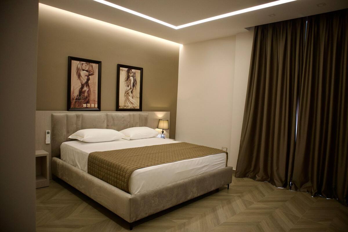 Quarto de hotel com uma cama grande, duas pinturas penduradas na parede, uma luminária de cabeceira e uma janela com cortinas.