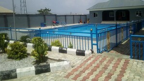 Uma piscina com grade azul, um edifício com telhado cinzento e um caminho pavimentado.