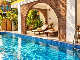 Piscina com borda azulejada, cercada por um arco de tijolos, e duas cadeiras de descanso com toalhas.