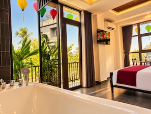 Quarto de hotel com uma banheira grande, uma cama com cobertor vermelho e vista para palmeiras e um edifício lá fora.