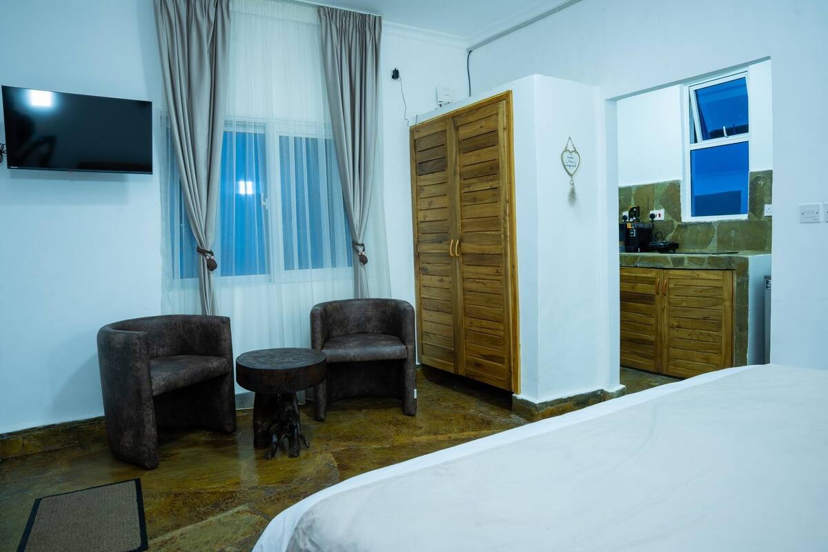 Quarto de hotel com cama, duas poltronas, uma mesa pequena, televisão, guarda-roupa e cozinha compacta.