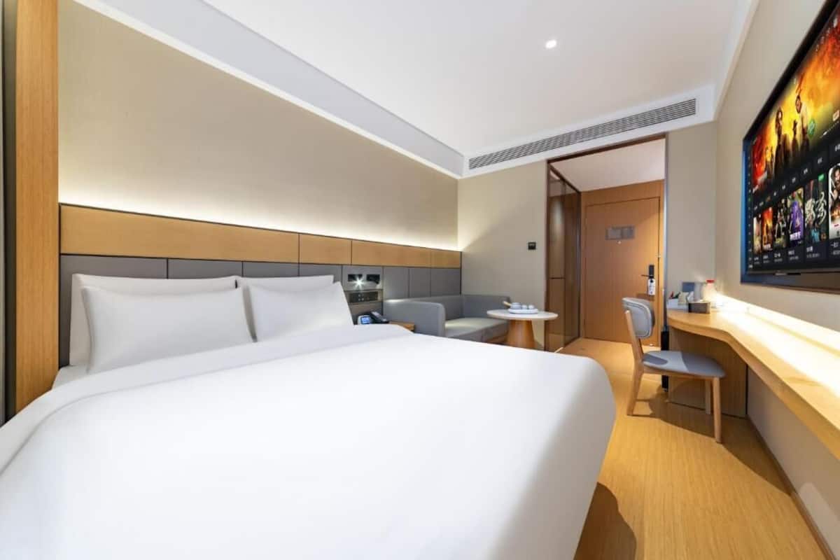 Quarto de hotel moderno com uma cama grande, uma escrivaninha com cadeira, uma mesinha e uma televisão na parede.