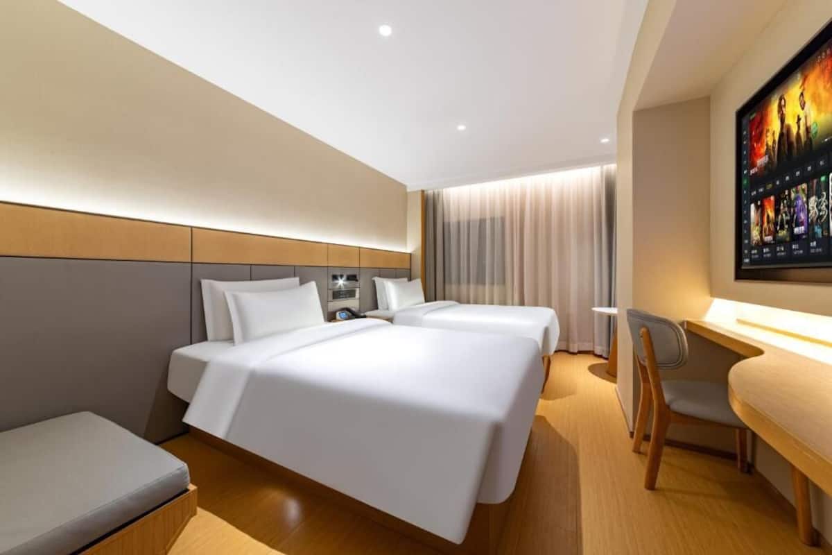 Quarto de hotel com duas camas, uma mesa, uma cadeira e uma televisão exibindo cartazes de filmes.