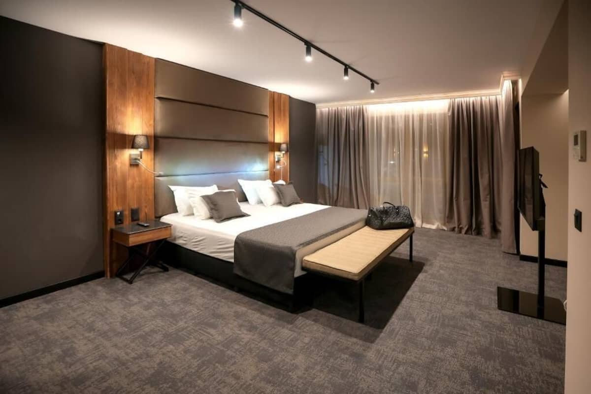 Quarto de hotel moderno com uma cama grande, mesas de cabeceira, um banco e vista para a cidade através das cortinas.