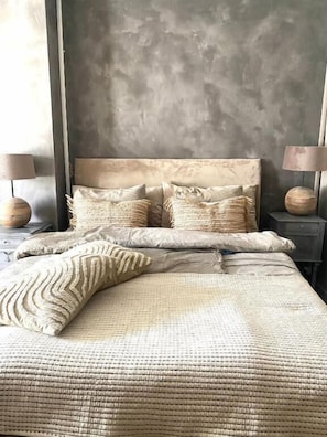 Cama com manta bege texturizada e almofadas, painel de cabeceira de madeira e duas luminárias laterais.