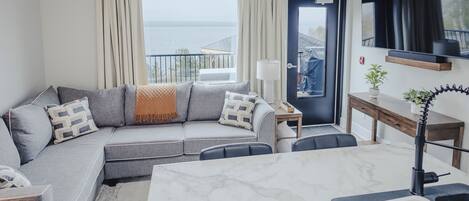 Uma sala de estar moderna com um sofá modular cinza, uma mesa de jantar de mármore, uma pequena mesa lateral e vista para o mar através de amplas janelas.