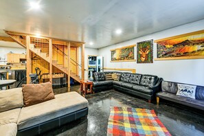 Uma sala de estar ampla com um sofá de couro preto, um sofá bege e um tapete colorido. Há uma escada que leva a um andar superior, e obras de arte estão expostas nas paredes.