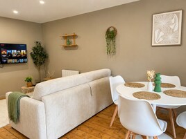 Uma sala de estar com sofá, mesa de jantar com cadeiras, televisão na parede e uma planta no chão.