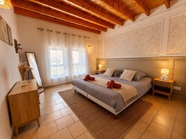 Quarto com cama, mesas de cabeceira, espelho, cómoda de madeira e janela com cortinas leves.