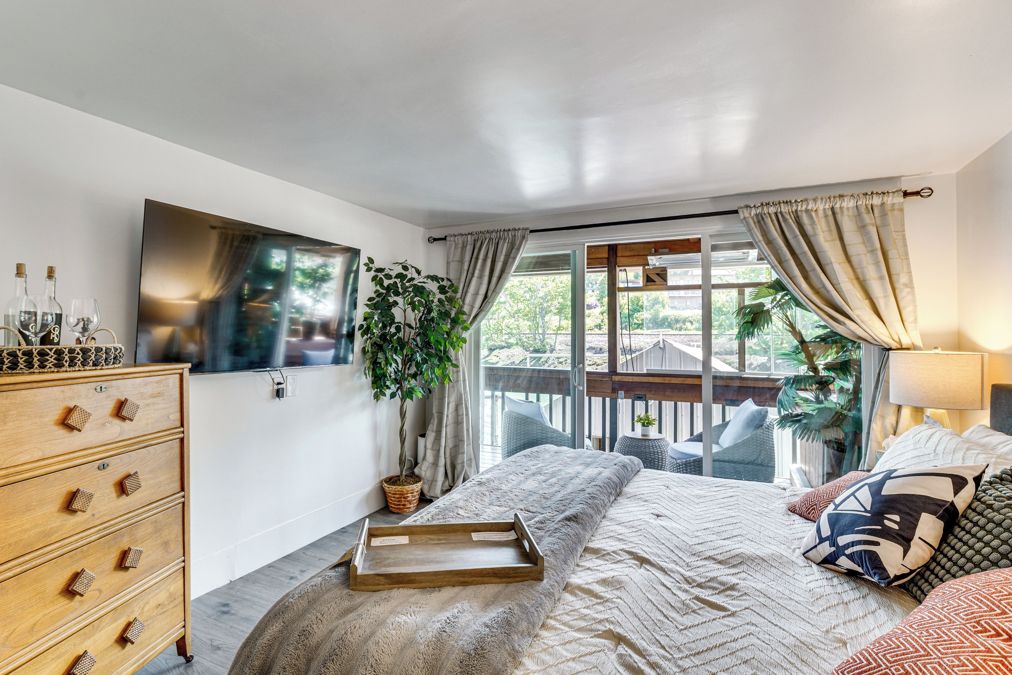 Quarto com cama, cômoda de madeira, televisão, planta e varanda com vista.