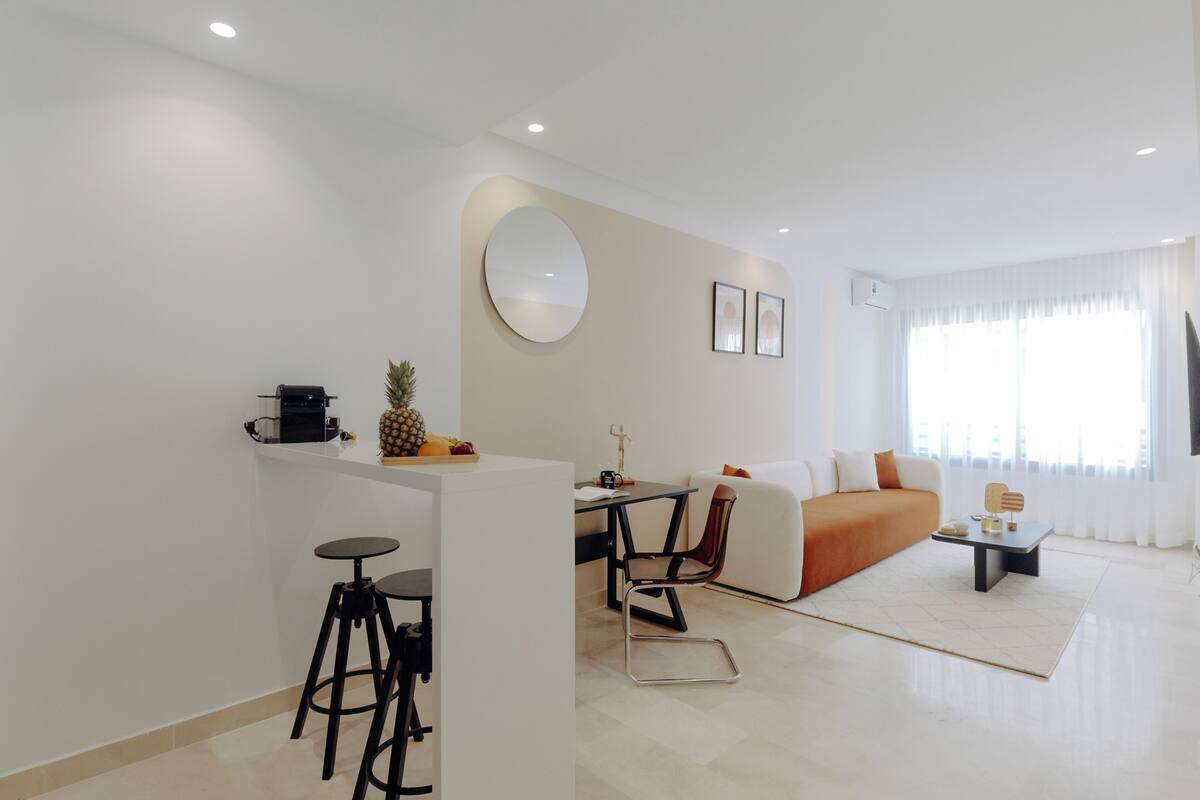 Uma sala moderna com sofá, mesa de centro, área de jantar com mesa e cadeiras, e bancada na cozinha com uma fruteira.