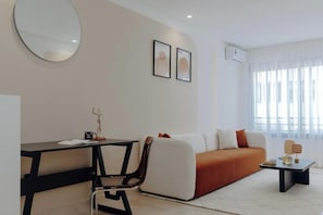 Quarto de hotel moderno com sofá, mesa de jantar, cadeira, uma pequena mesa com objeto decorativo e um espelho grande na parede.