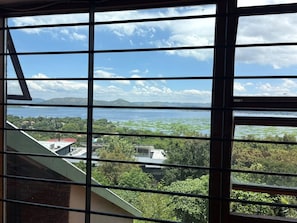 Vista pela janela, com padrão de grade, sobre um corpo de água, vegetação e montanhas ao longe.