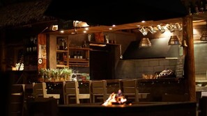 Uma área de bar ao ar livre com móveis de madeira, uma lareira e luzes penduradas.